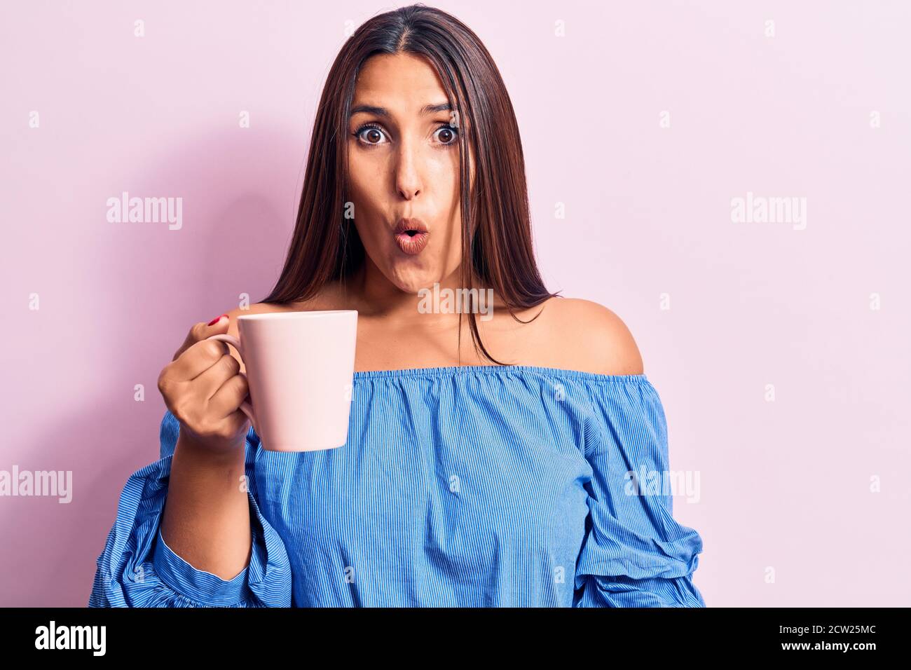 Young beautiful brunette woman drinking mug of coffee scared and amazed ...