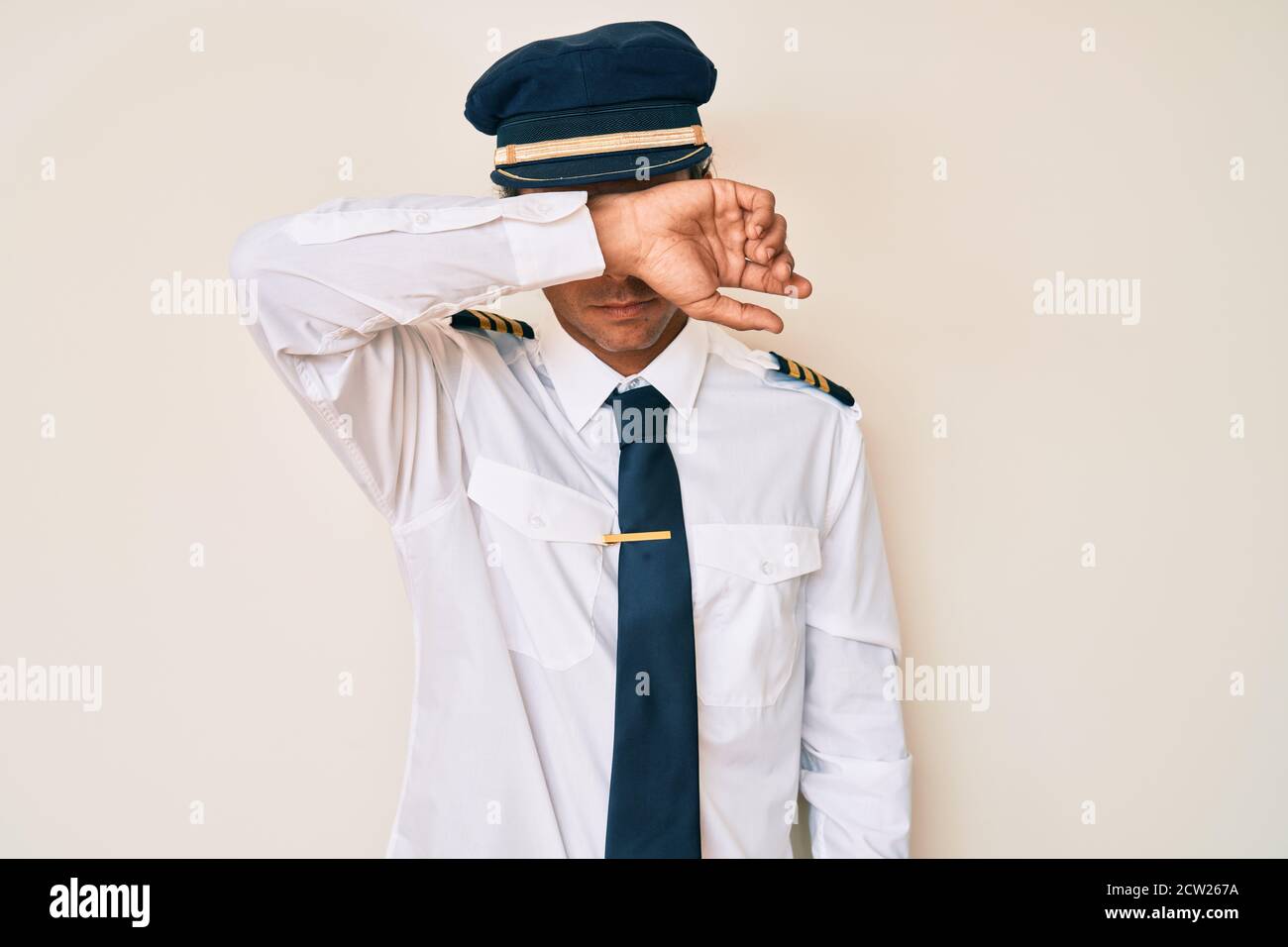 Young hispanic man wearing airplane pilot uniform covering eyes with ...