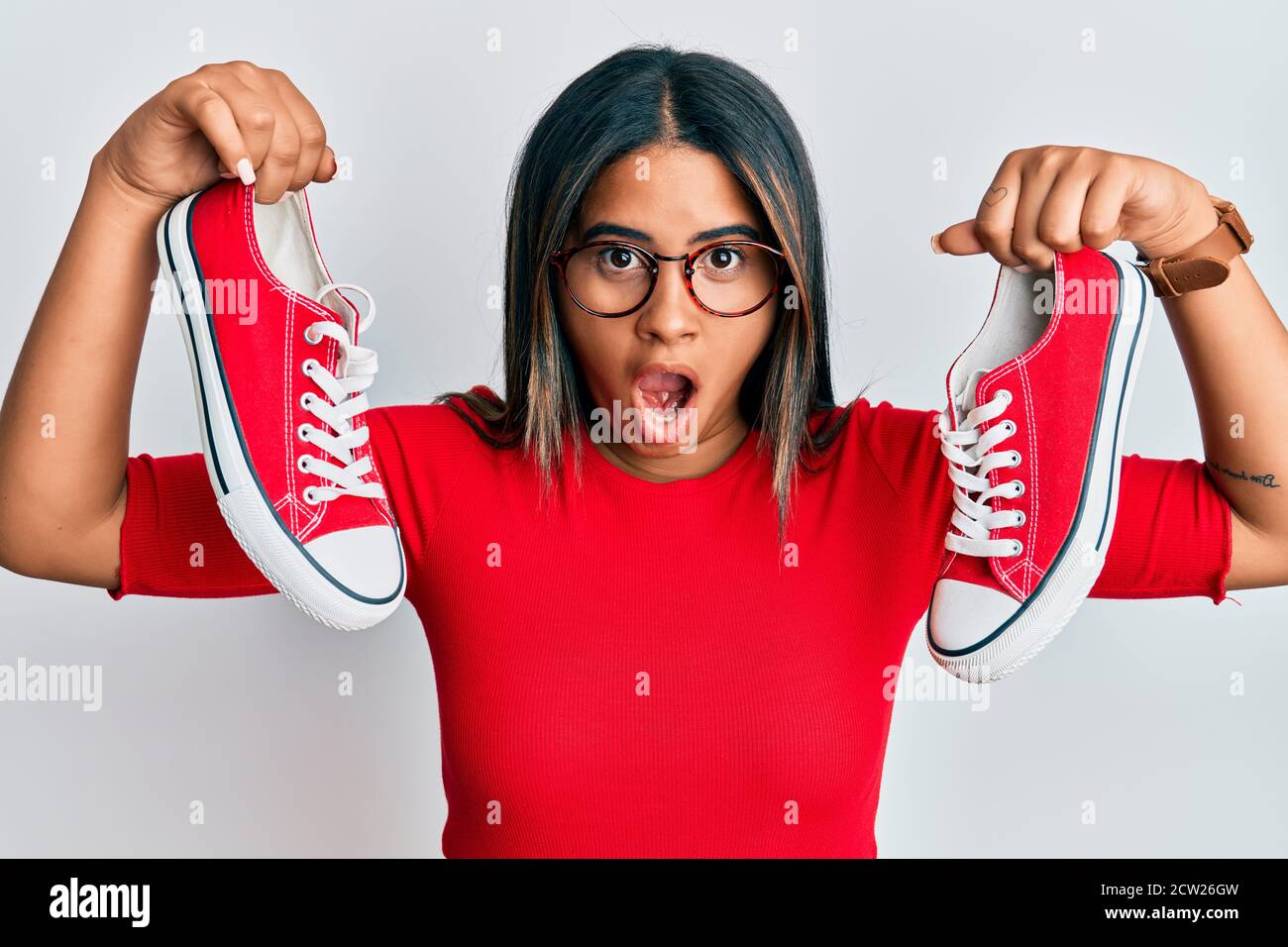 Young latin girl holding red casual shoes afraid and shocked with ...