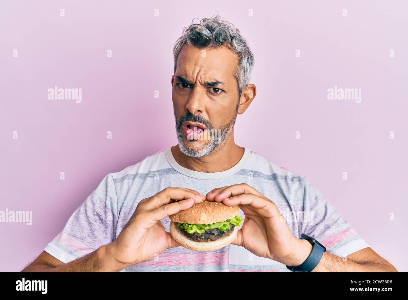 Middle age grey-haired man eating a tasty classic burger in shock face ...