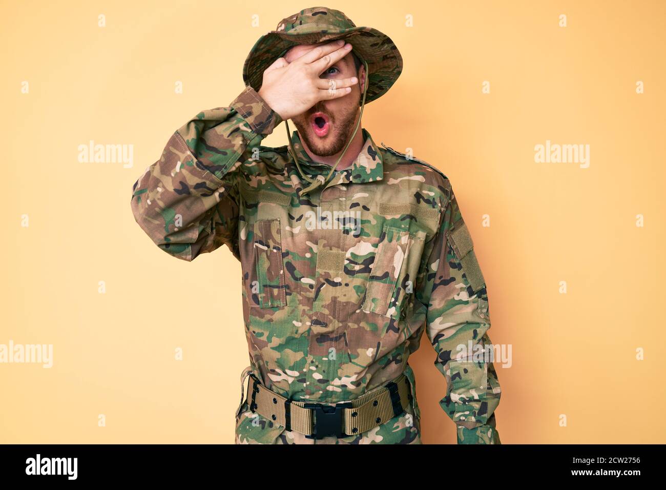 Young caucasian man wearing camouflage army uniform peeking in shock ...