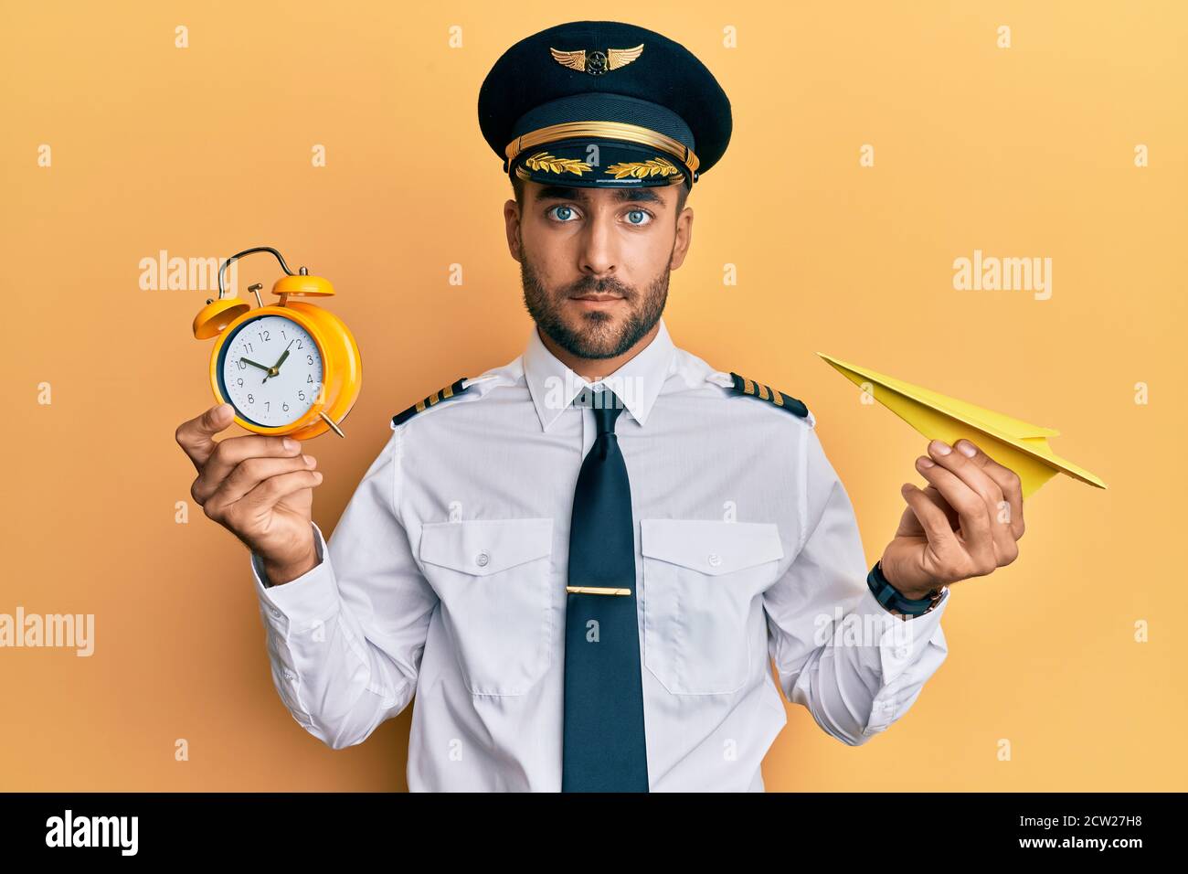 Handsome hispanic pilot man holding paper plane and alarm clock relaxed ...