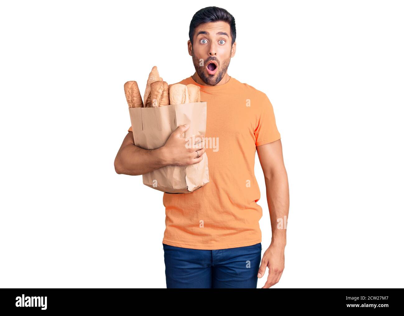 Young hispanic man holding paper bag with bread scared and amazed with ...