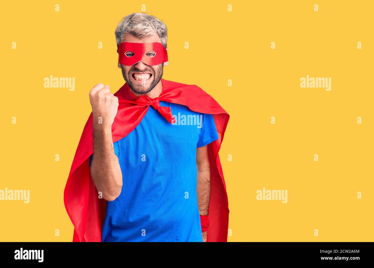 Young blond man wearing super hero custome angry and mad raising fist ...