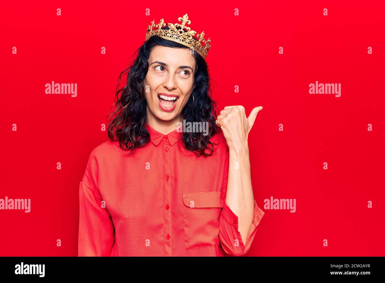 Young beautiful hispanic woman wearing princess crown pointing thumb up ...