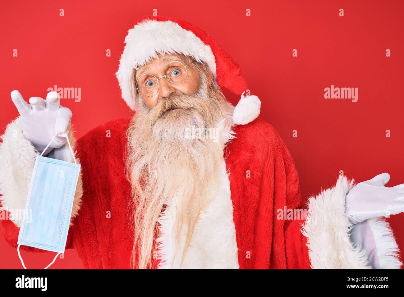 Old senior man wearing santa claus costume holding safety mask ...