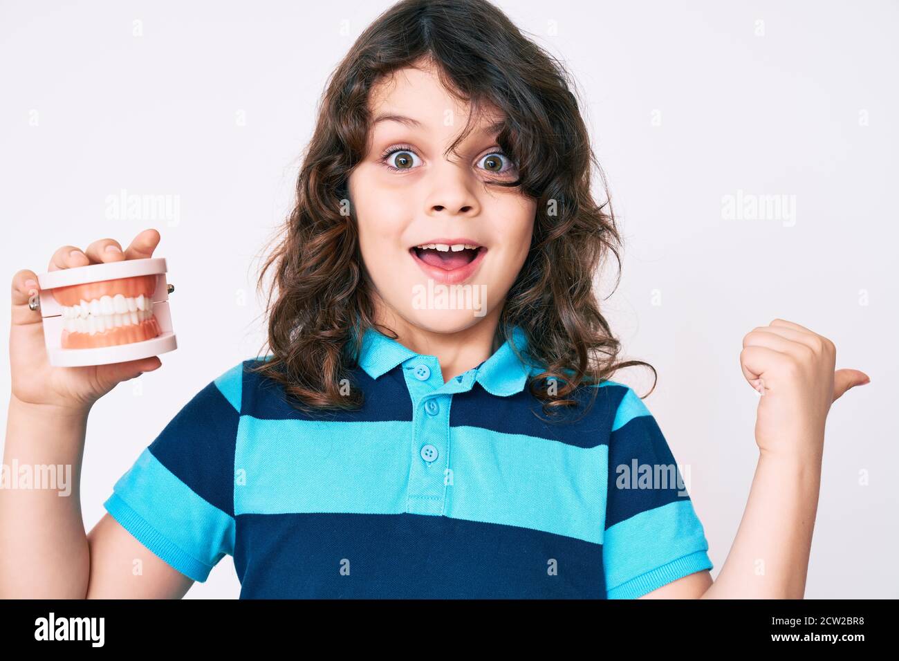 Cute hispanic child with long hair holding denture pointing thumb up to ...
