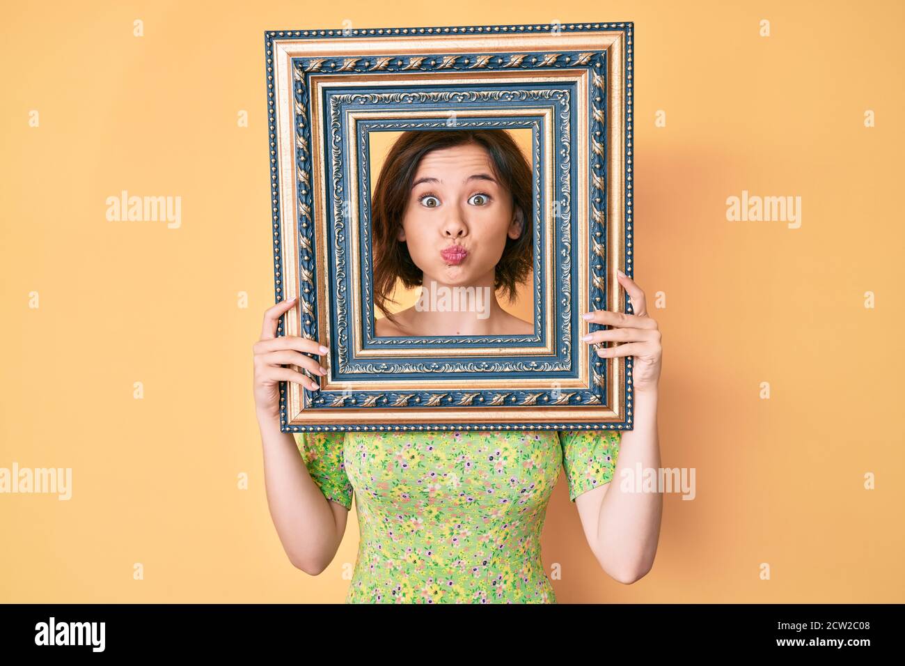 Young beautiful woman holding empty frame puffing cheeks with funny ...