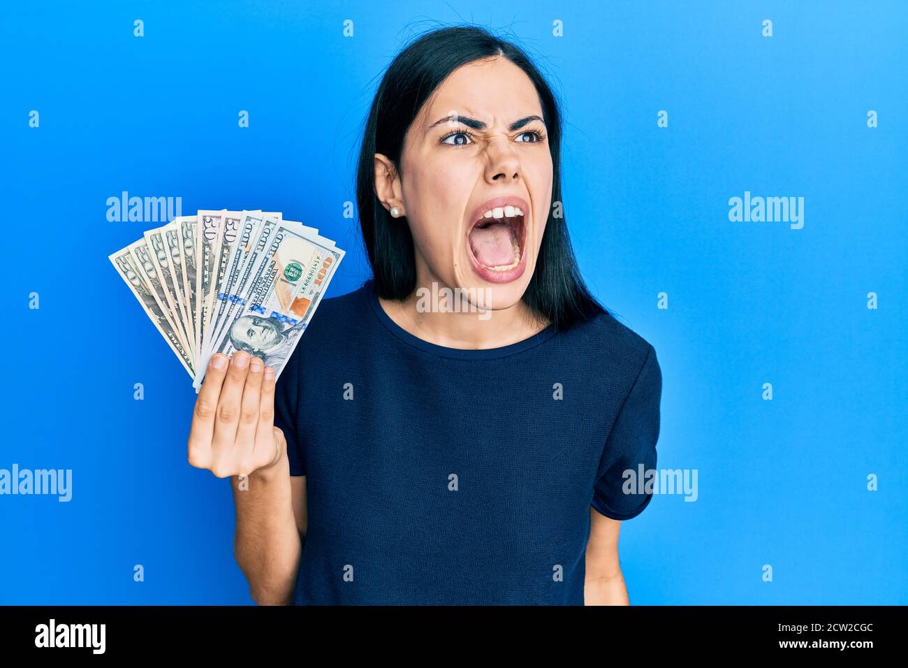 Beautiful young woman holding dollars angry and mad screaming ...