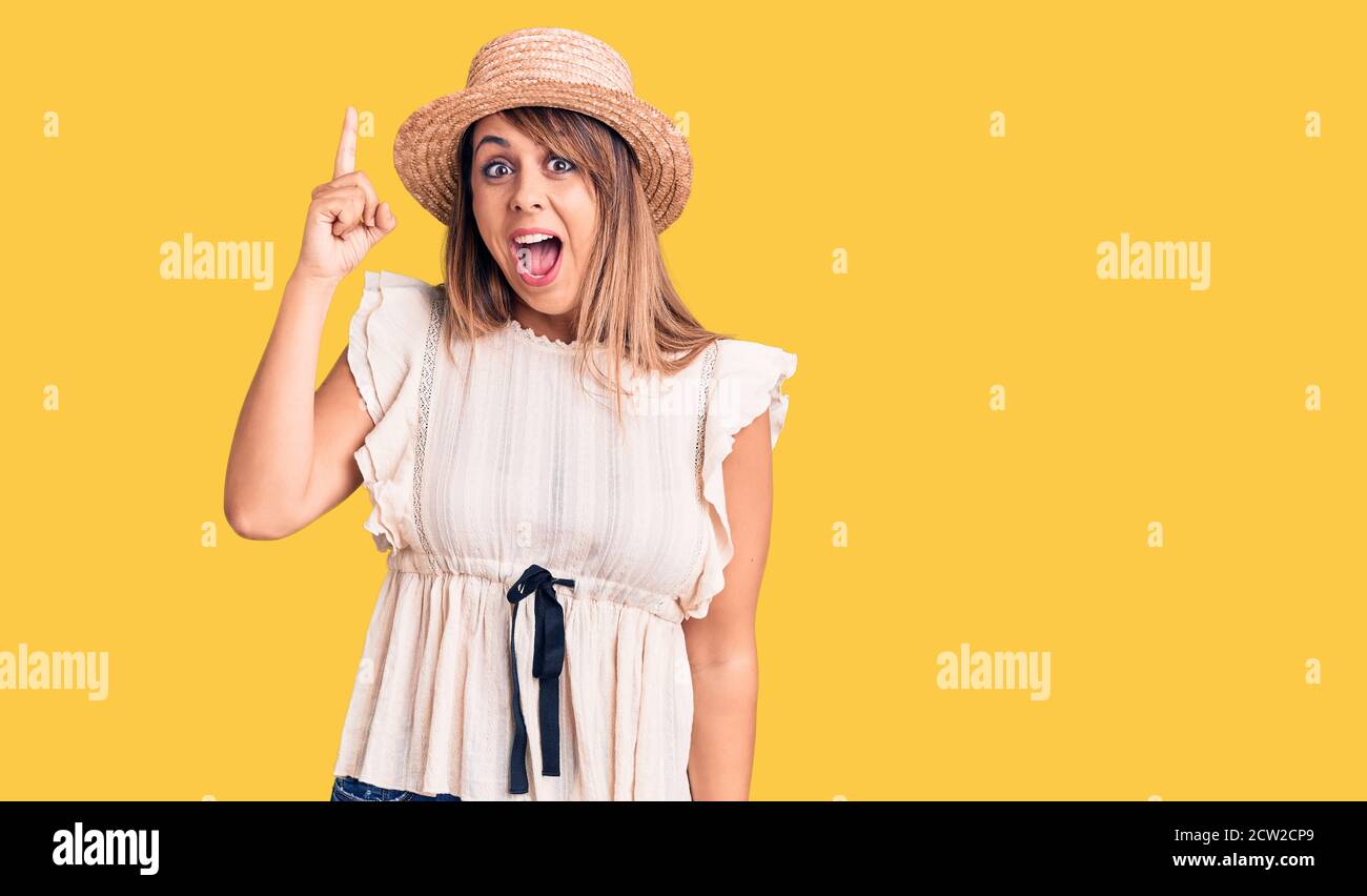 Young beautiful woman wearing summer hat and t-shirt pointing finger up ...