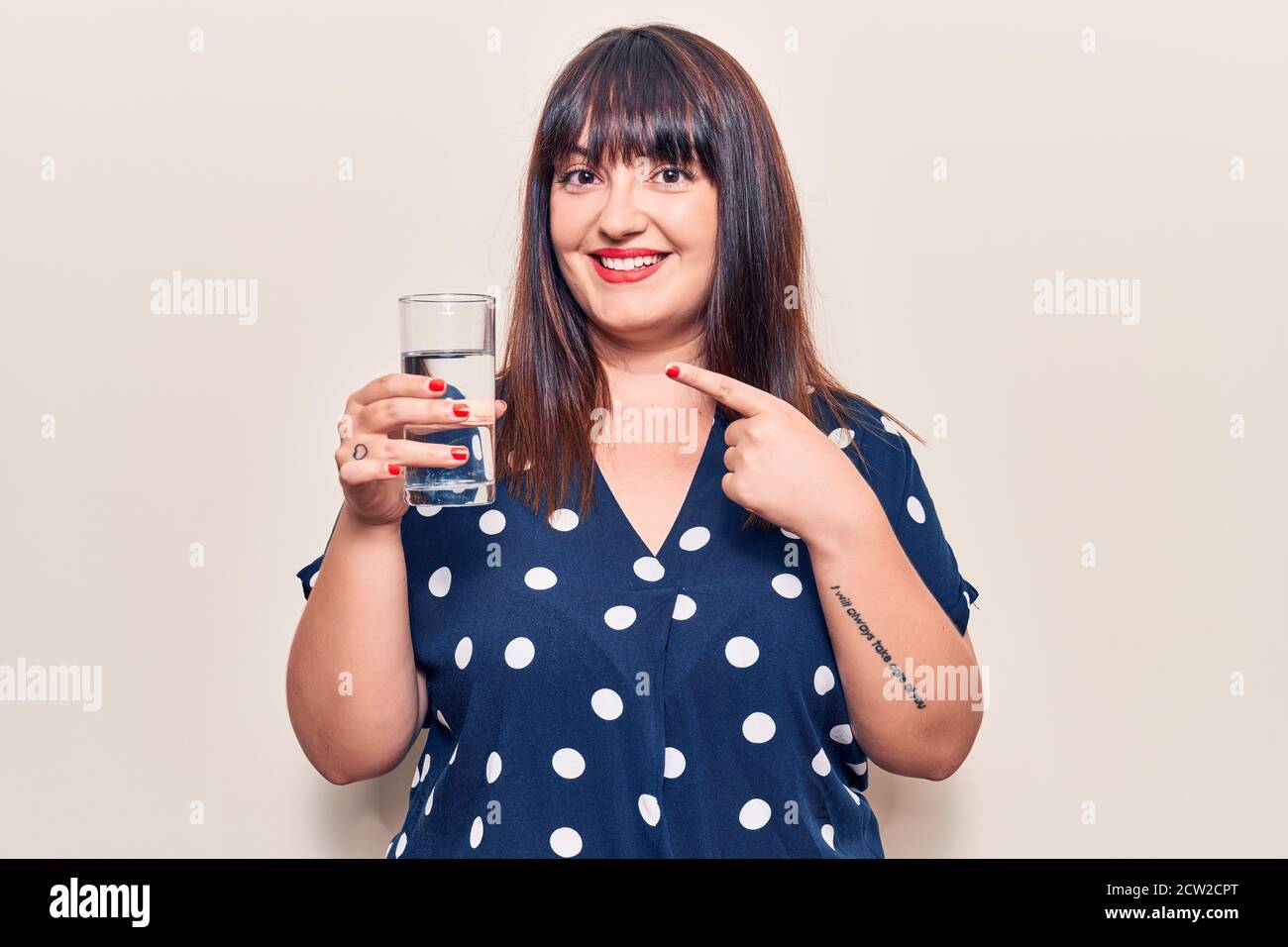Young plus size woman drinking glass of water smiling happy pointing ...