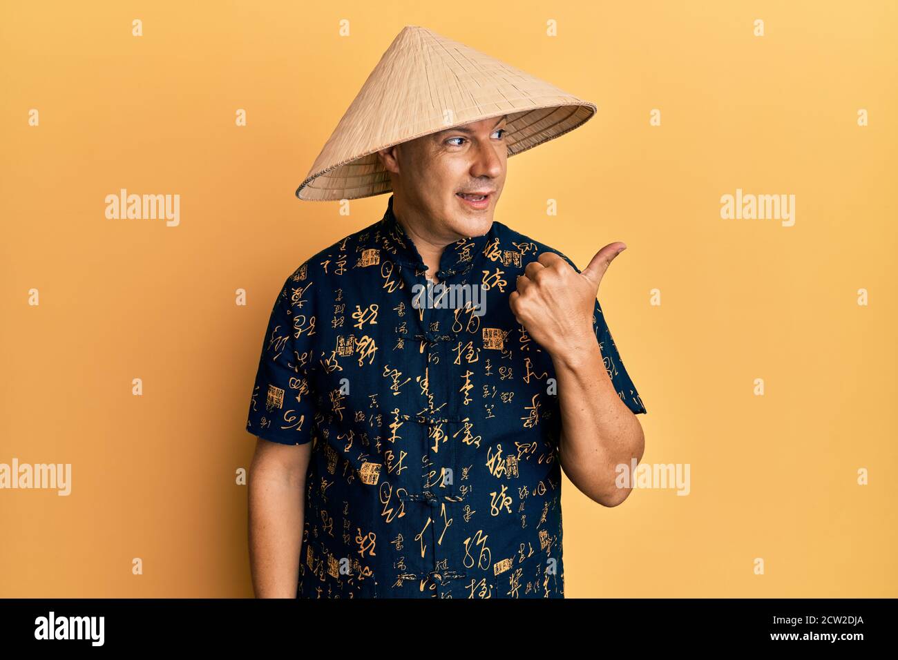 Middle age bald man wearing traditional asian straw hat pointing thumb ...