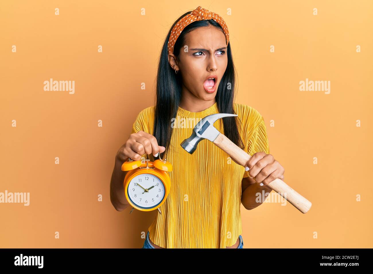 Young brunette woman holding alarm clock and hammer angry and mad ...