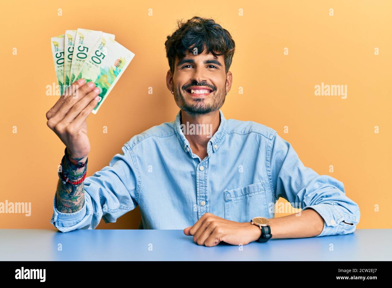 Young hispanic man holding israel shekels sitting on the table looking ...