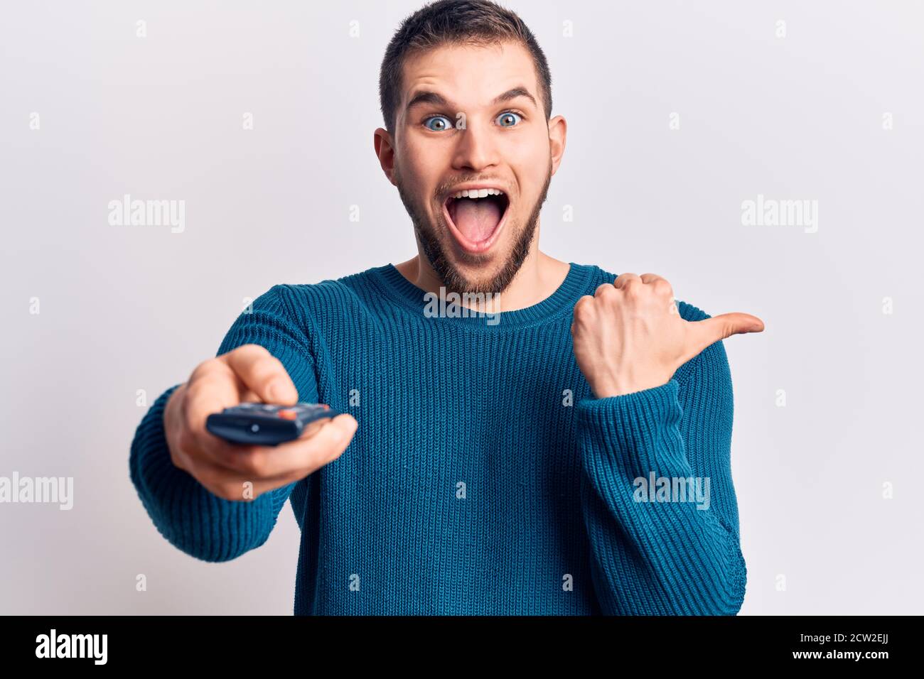 Young handsome man holding tv remote control pointing thumb up to the ...