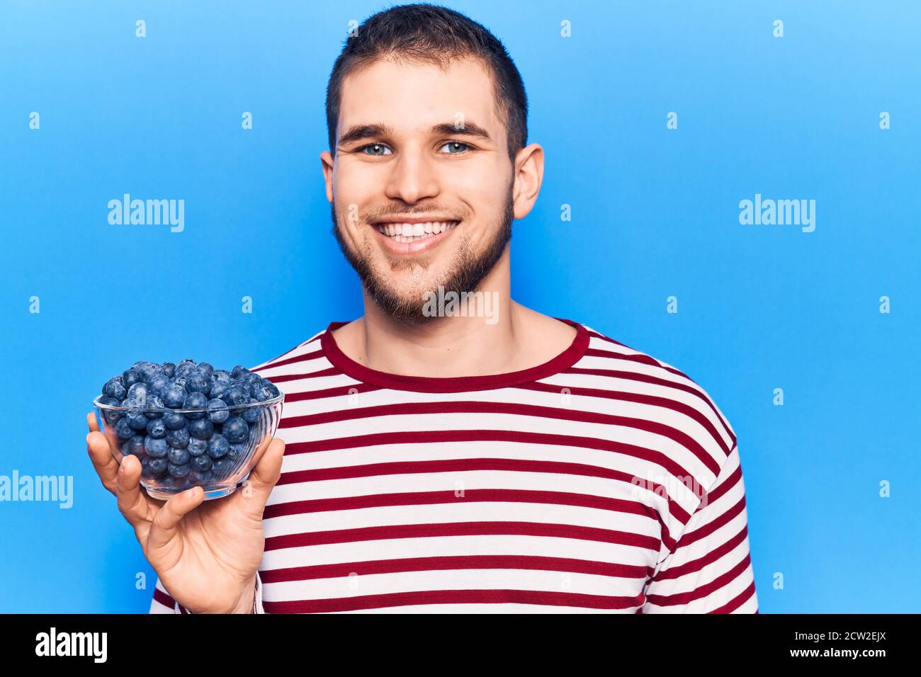 Young handsome man holding bowl with blueberries looking positive and ...