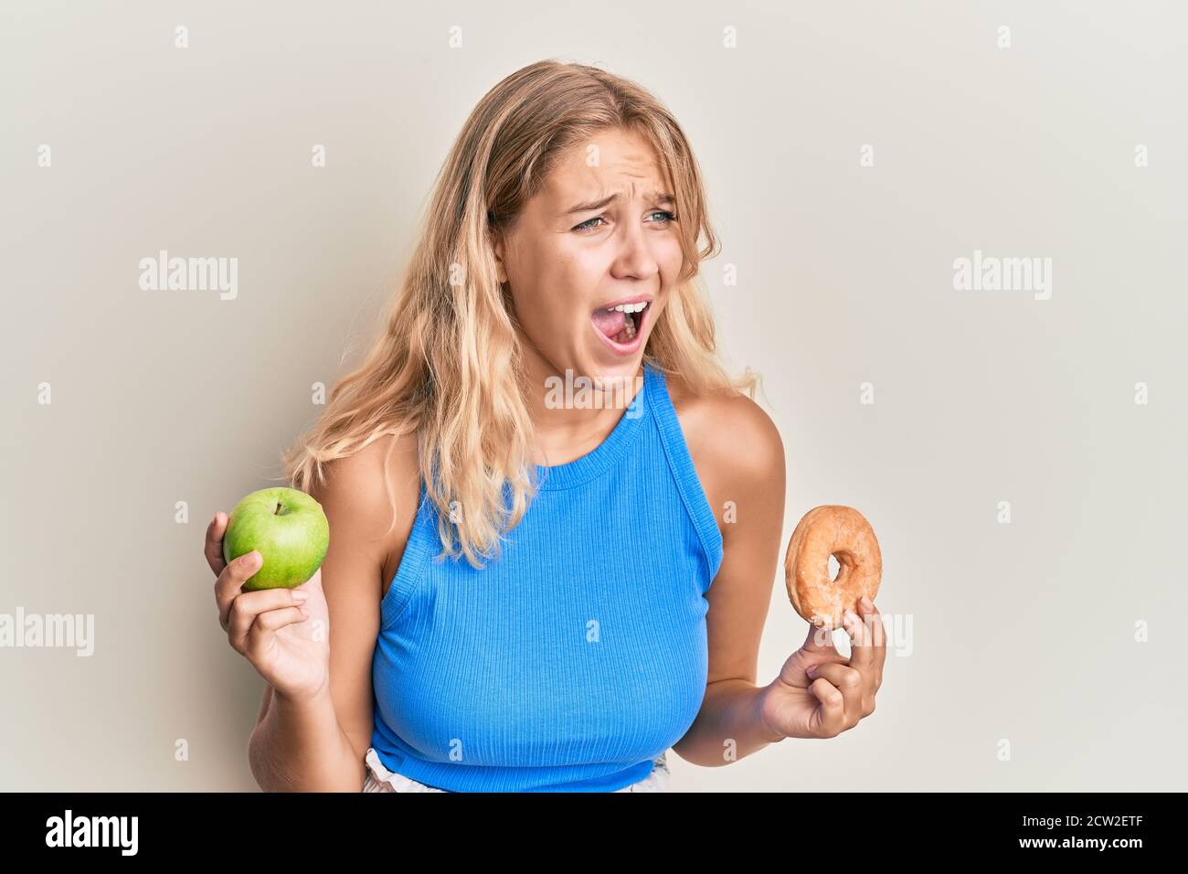 Young blonde girl holding green apple and donut angry and mad screaming ...