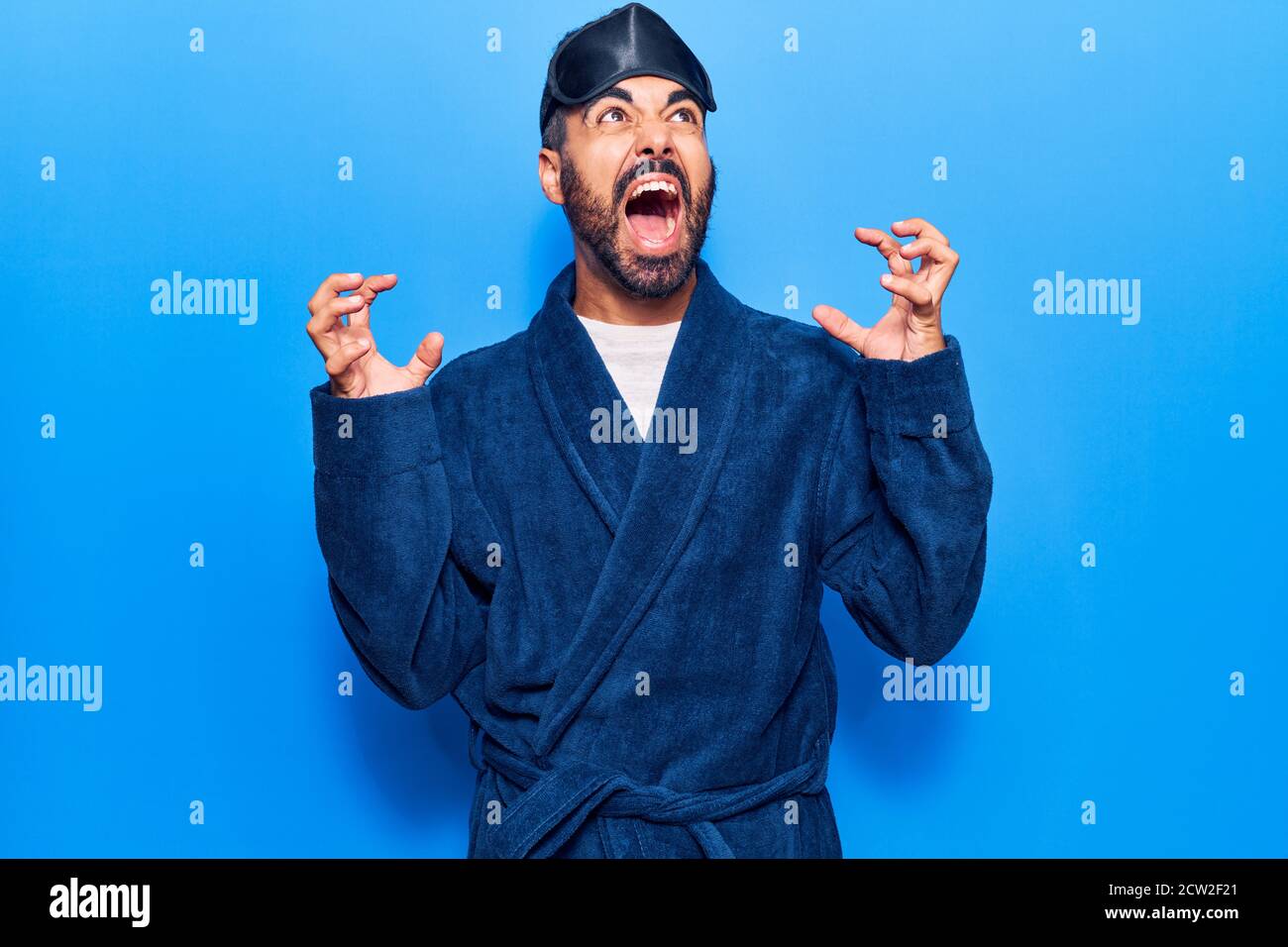 Young hispanic man wearing sleep mask and robe crazy and mad shouting ...