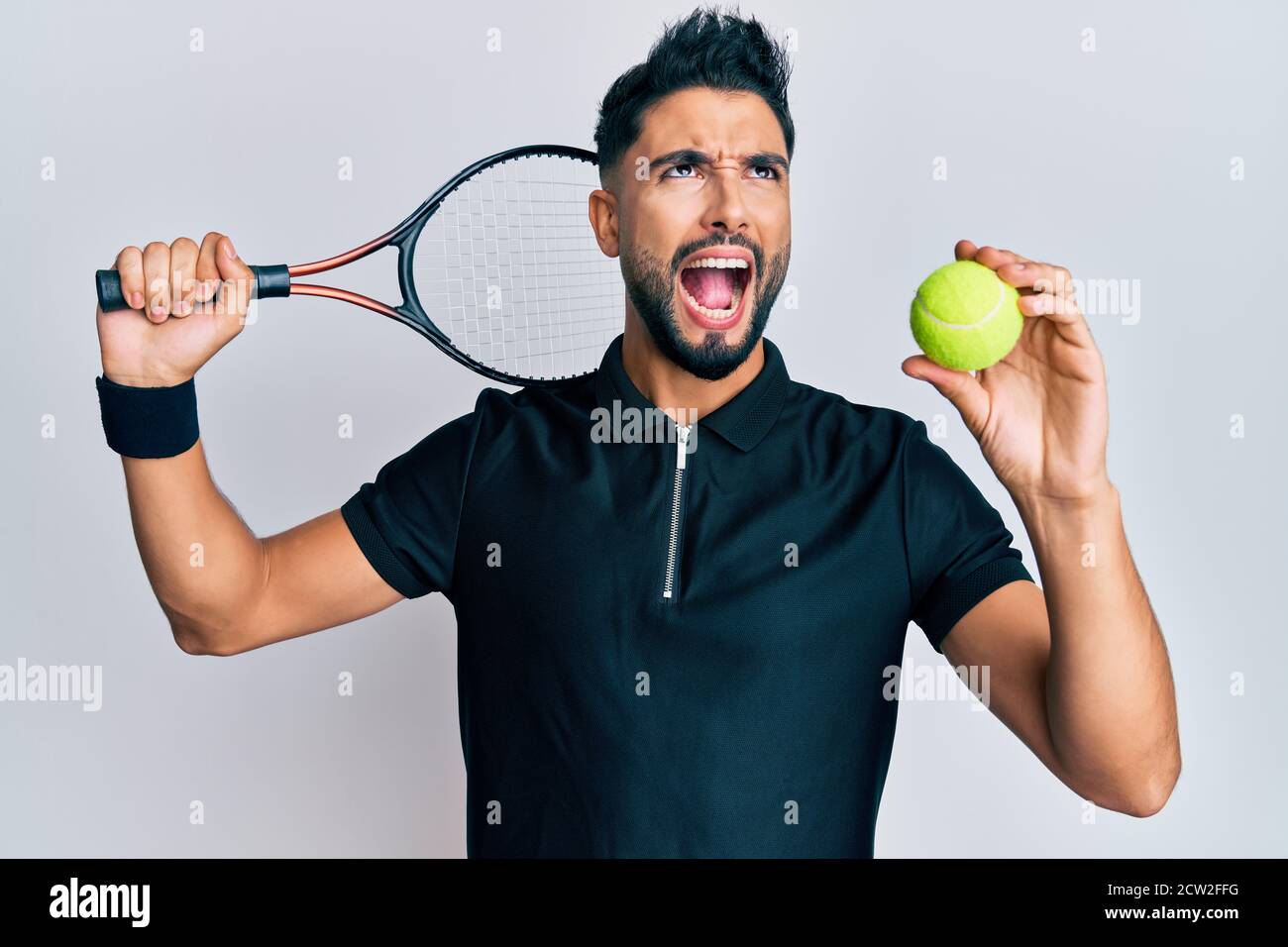 Young man with beard playing tennis holding racket and ball angry and ...