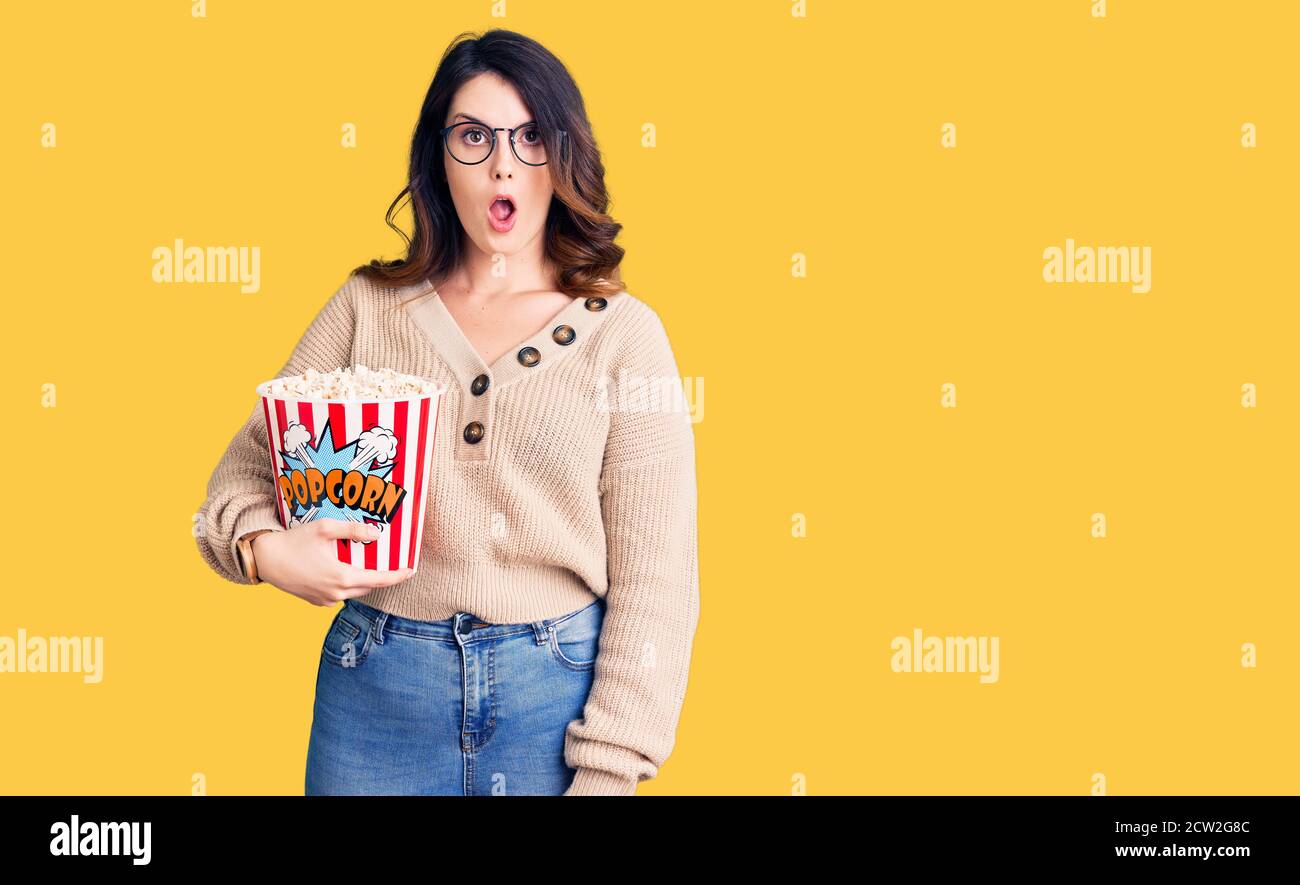 Beautiful young brunette woman eating popcorn scared and amazed with ...