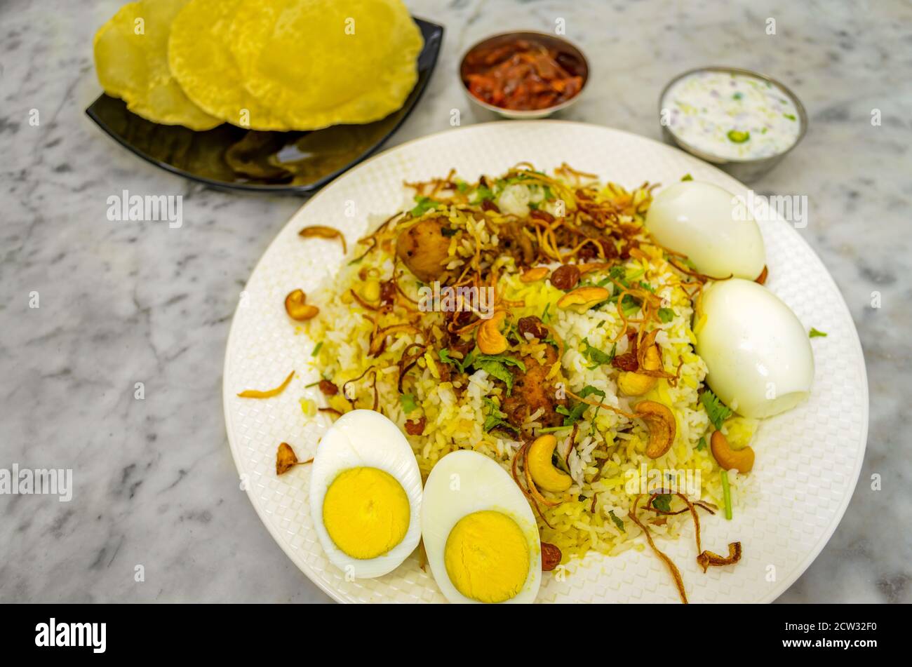 Chicken Biryani with eggs, papads, pickle and raita on a white marble ...
