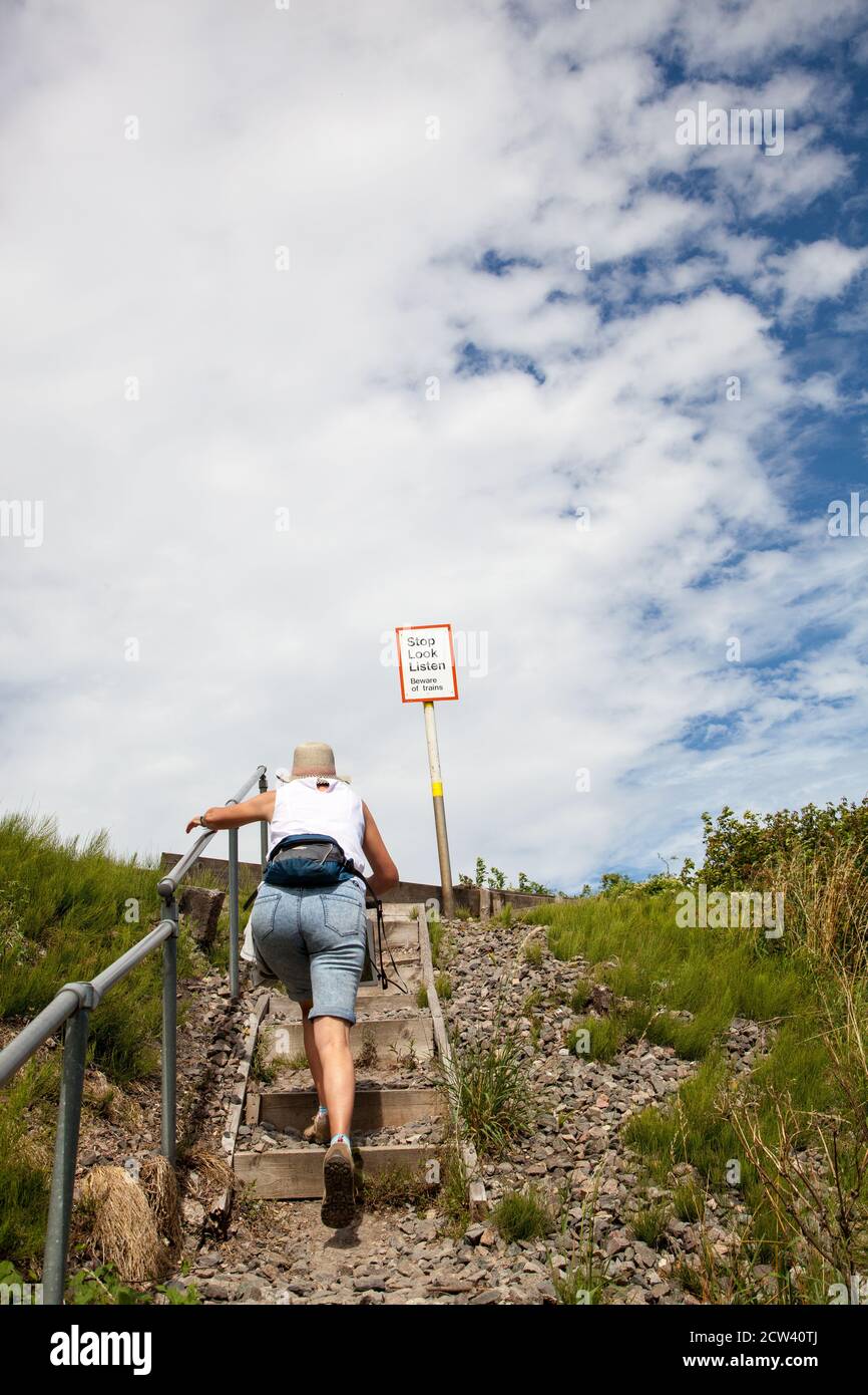 Woman walking up steps to a footpath to cross a railway track with sign ...