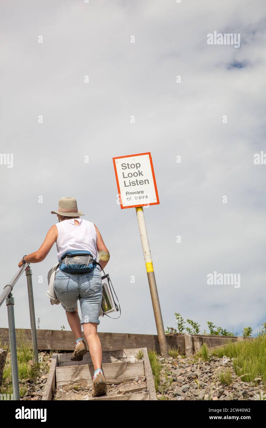 Woman walking up steps to a footpath to cross a railway track with sign ...