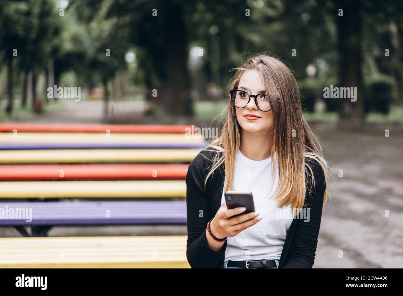 Young girl in glasses looks into a cellphone. Sitting on a park bench. Concept of communication ...