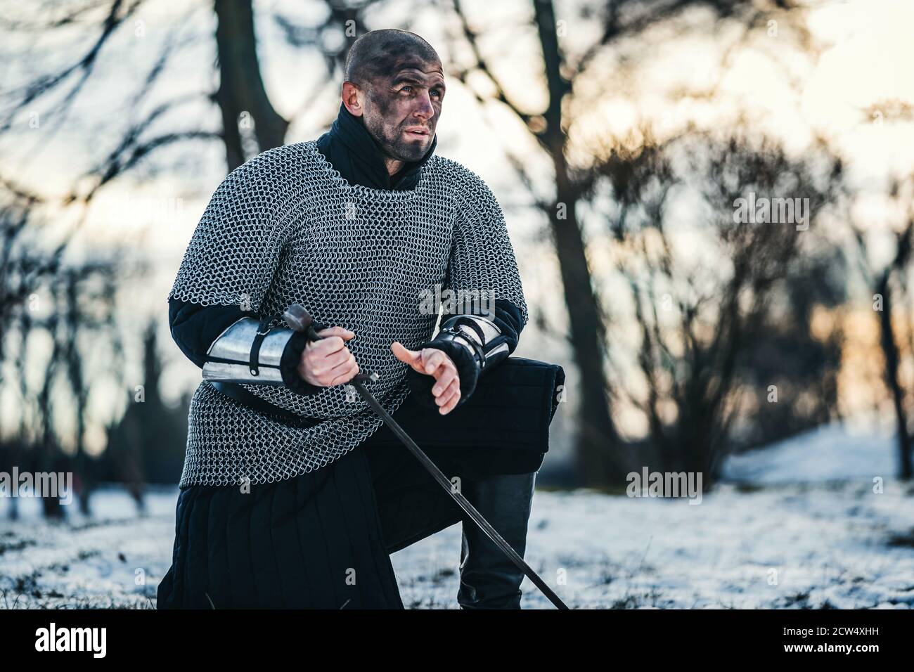 A medieval warrior in chain mail armor kneeling with his sword in his hand and dirty face after ...