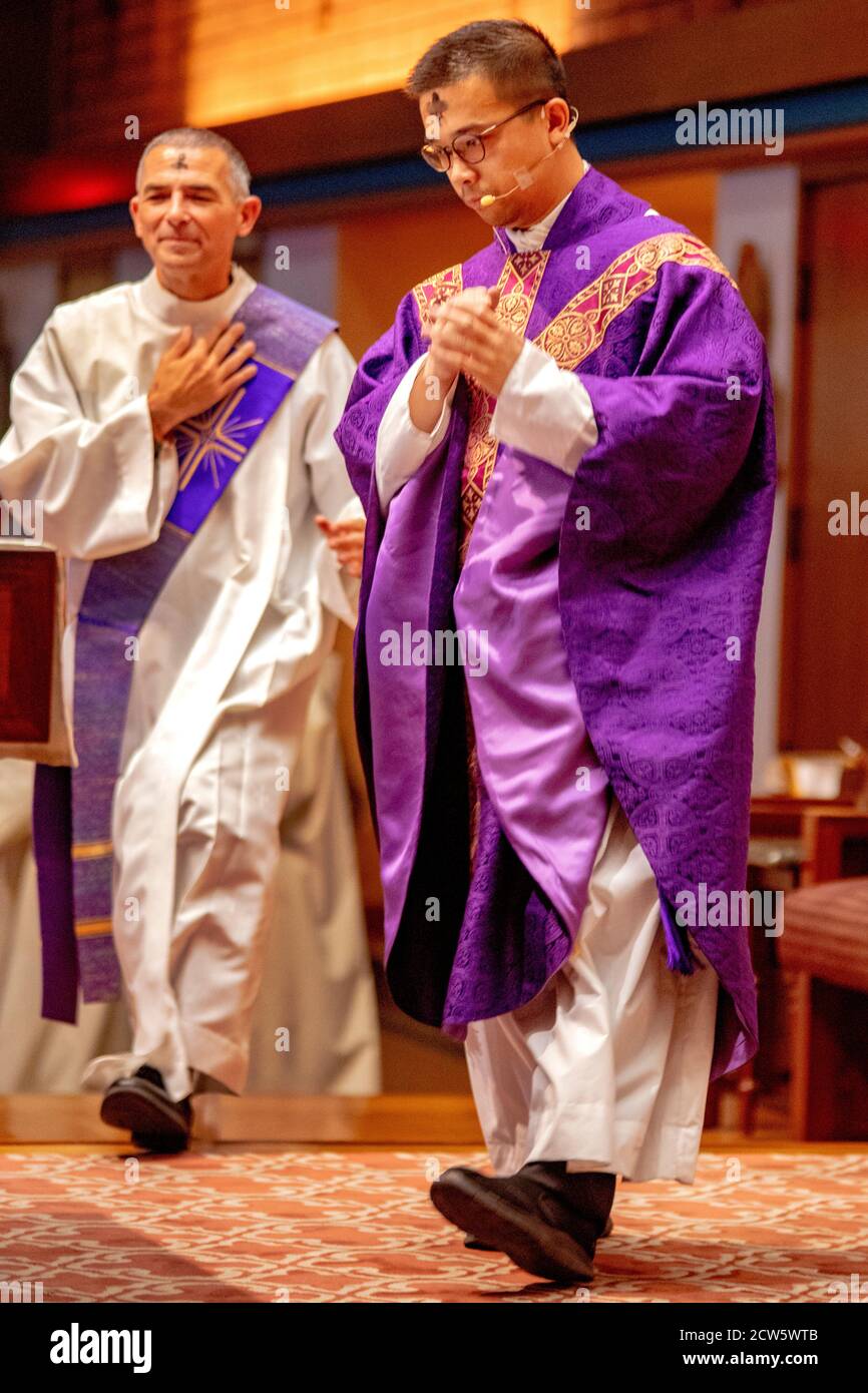 Wearing ceremonial robes, a deacon assists an Asian American priest ...