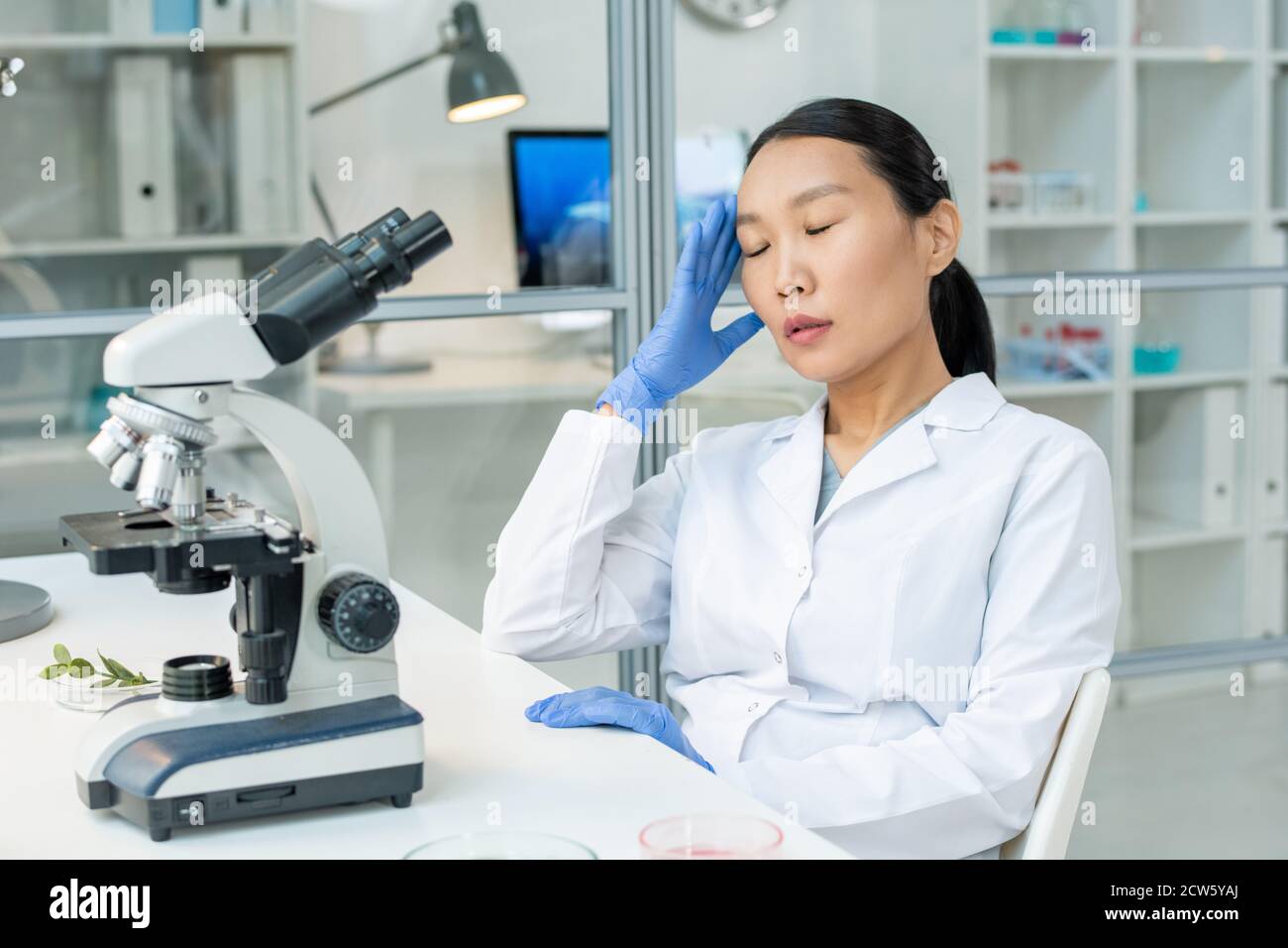 Young tired female laboratory worker in gloves and whitecoat touching ...