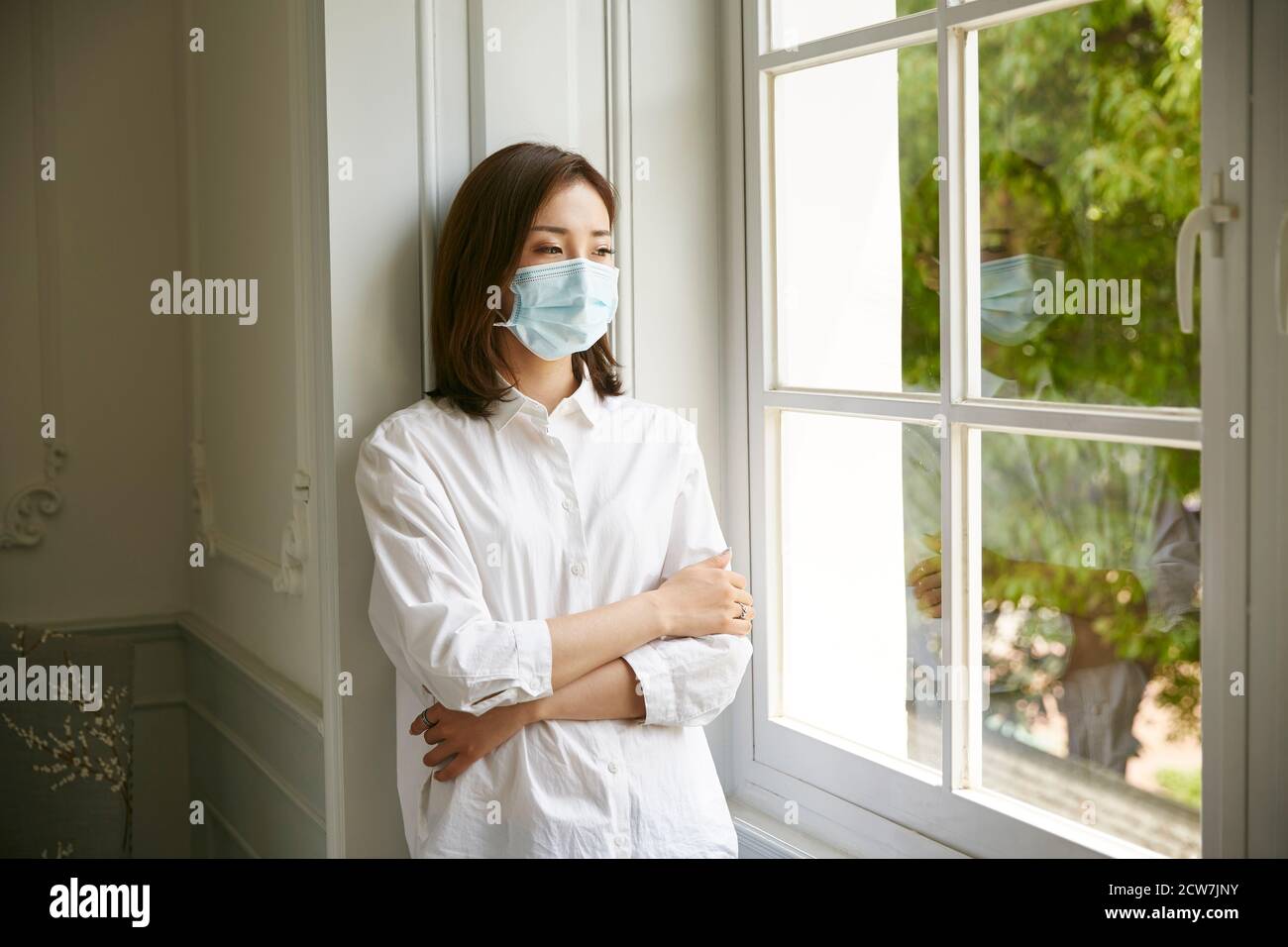 young asian woman in isolation wearing mask standing by window looking ...