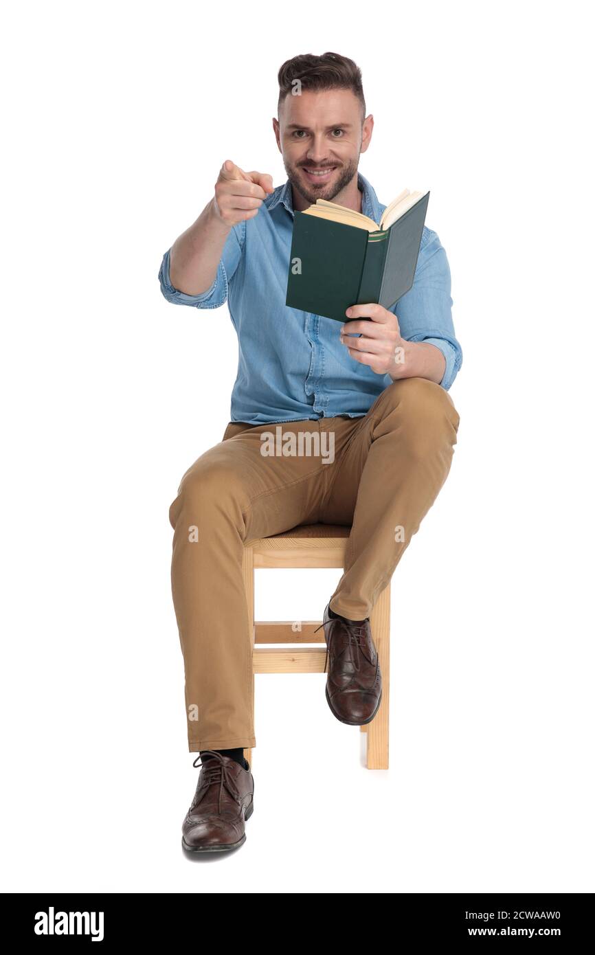 young casual man holding book and pointing finger, smiling and sitting ...