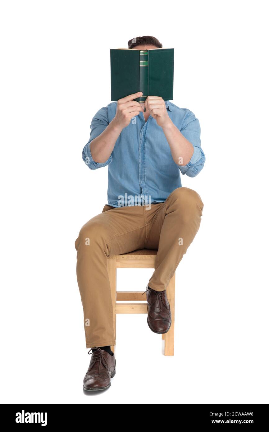 young casual man hiding behind book and sitting isolated on white ...