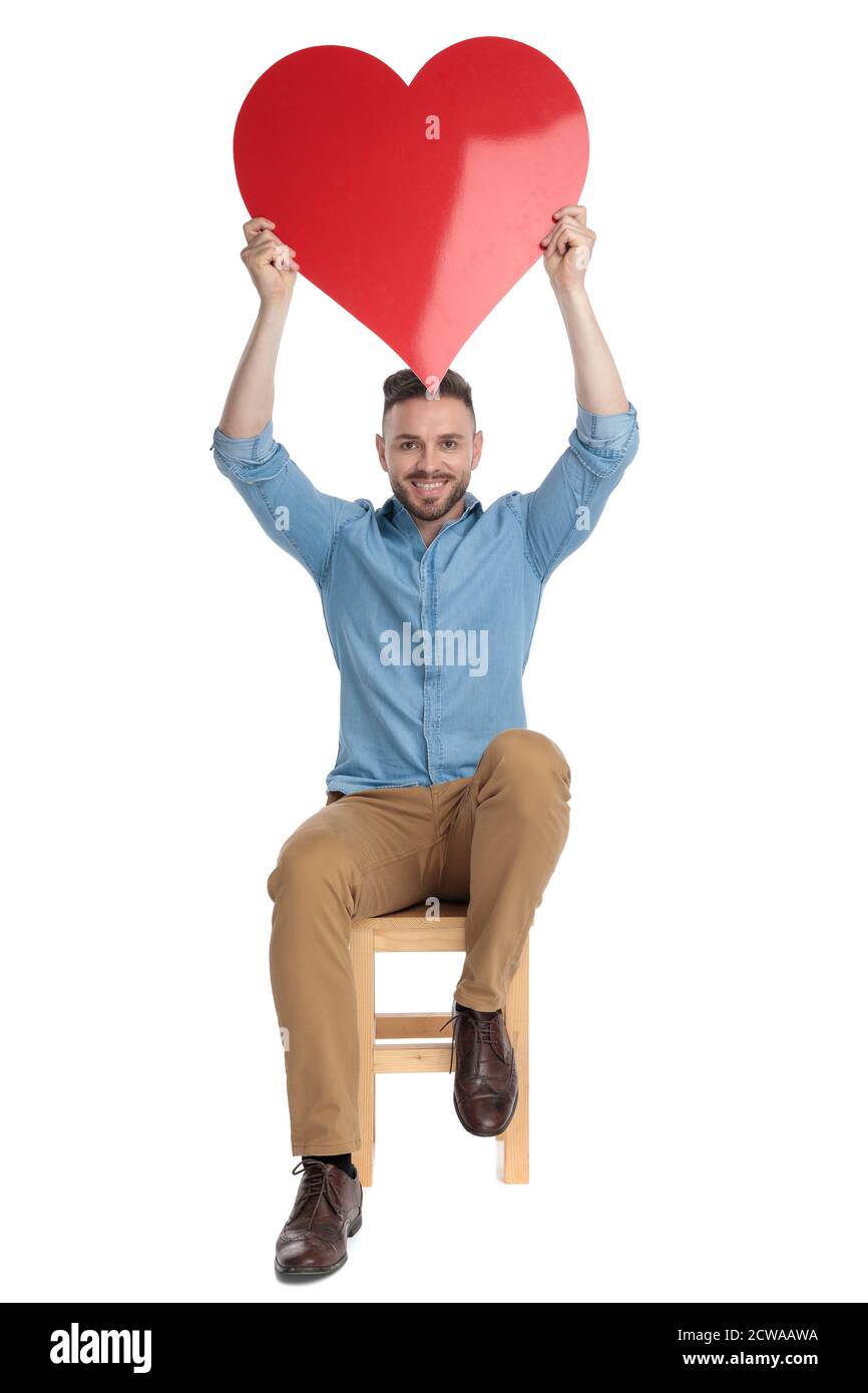 young casual man holding big red heart above head and smiling, sitting ...