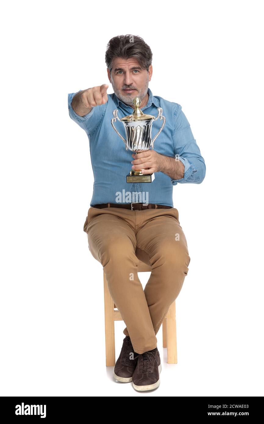 Positive casual man holding a trophy and pointing forward while sitting ...
