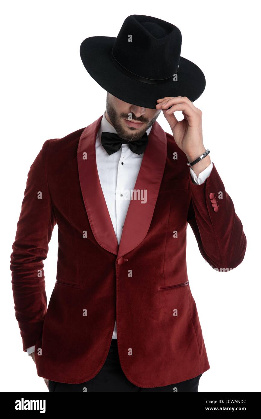 mysterious young man in red velvet tuxedo looking down and fixing hat ...