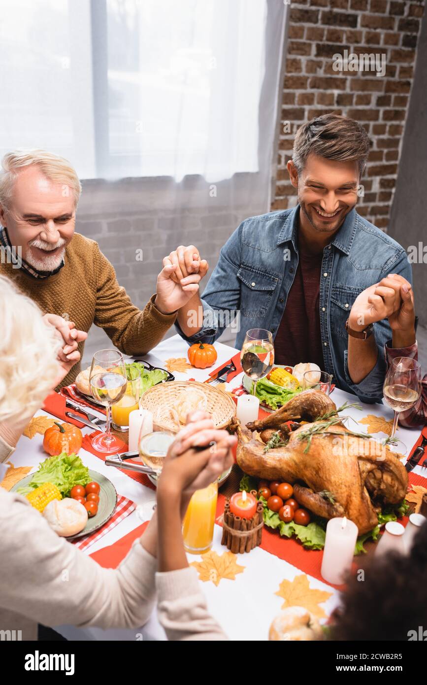 joyful multicultural family holding hands during dinner on thanksgiving ...