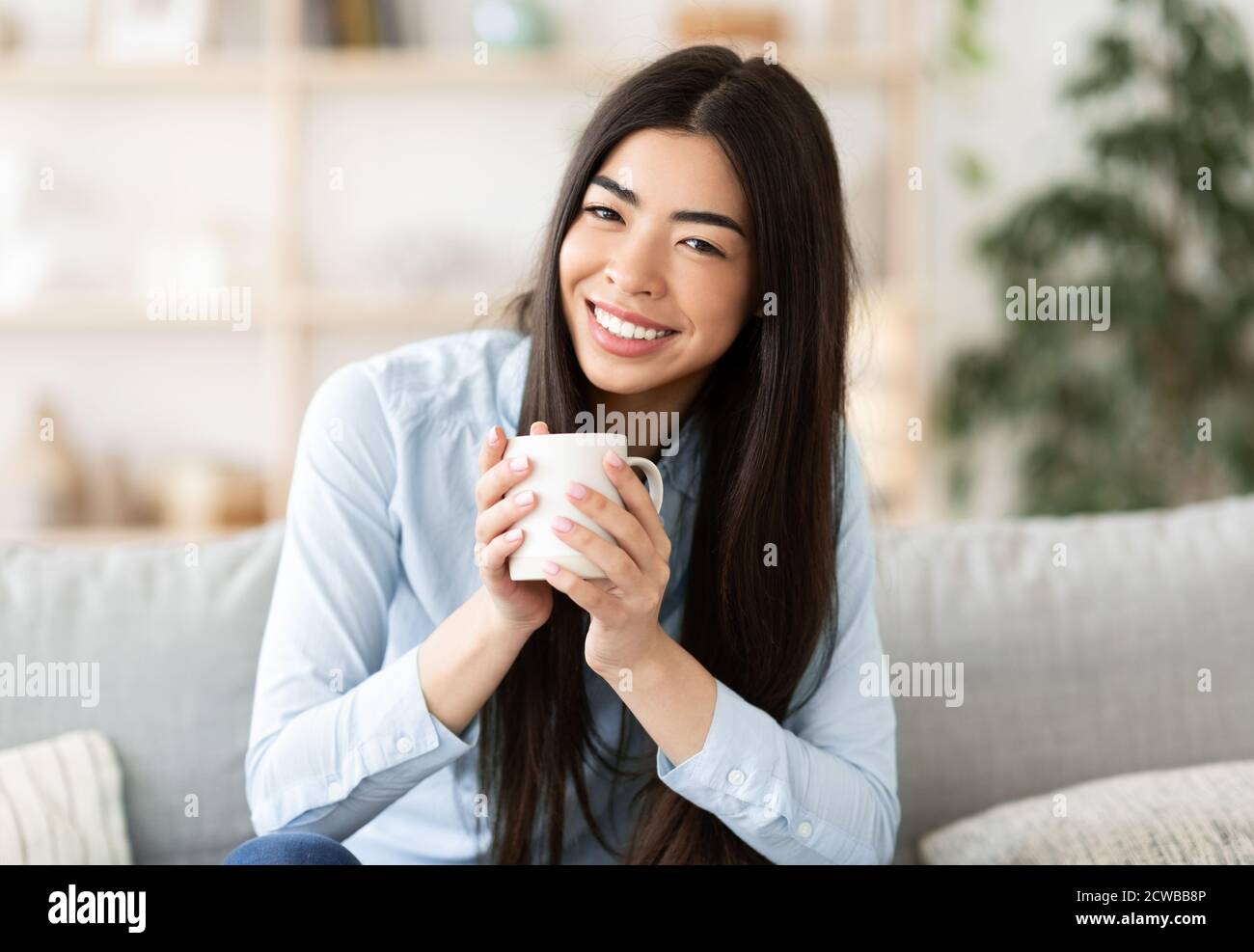 Smiling korean woman drinking coffee while resting on couch at home ...