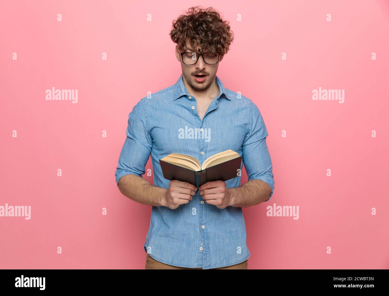 impressed young guy in denim shirt reading a book and standing on pink ...