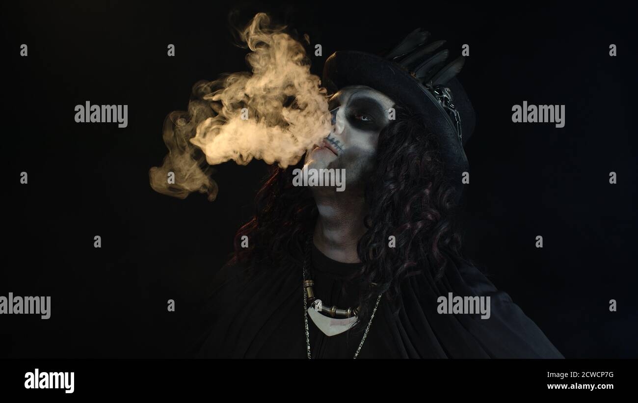 Sinister man with professional skull makeup exhaling cigarette smoke ...