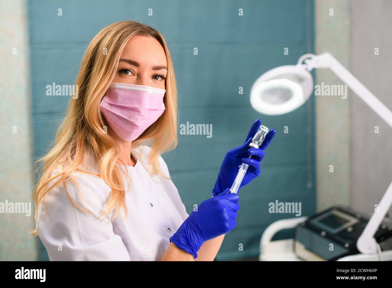Pretty blonde cosmetology doctor in pink disposable face mask and blue ...