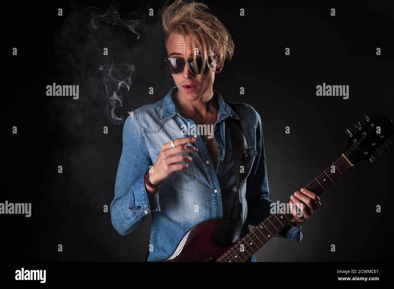 amazed young guitarist smoking cigarette in studio while holding guitar ...