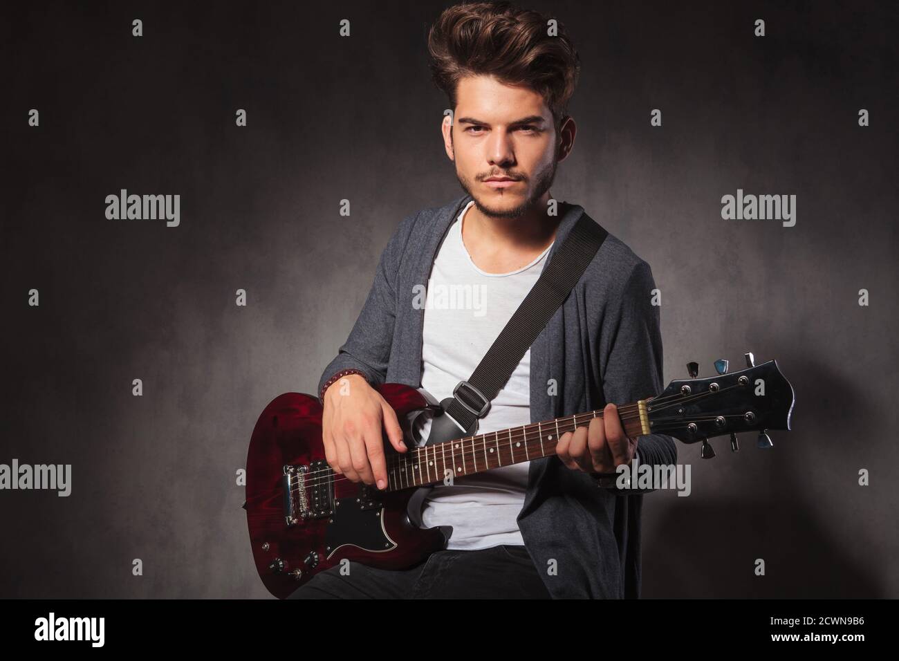 portrait of fashionable artist playing guitar while sitting in dark ...