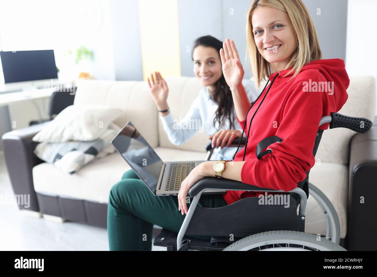 Young disabled woman in wheelchair with helper is sitting by laptop ...