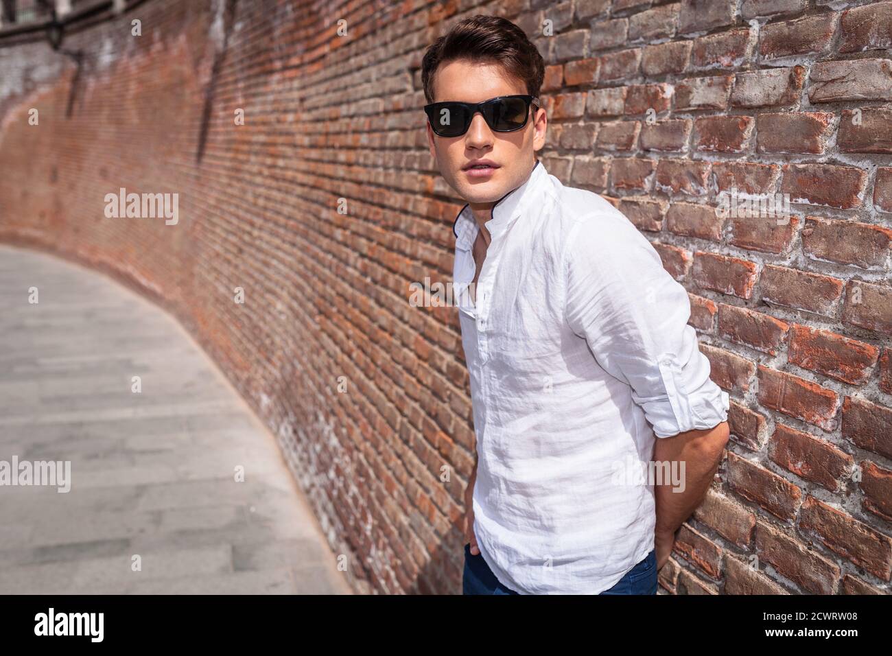 Side view of a casual fashion man leaning on a brick wall with his hand ...