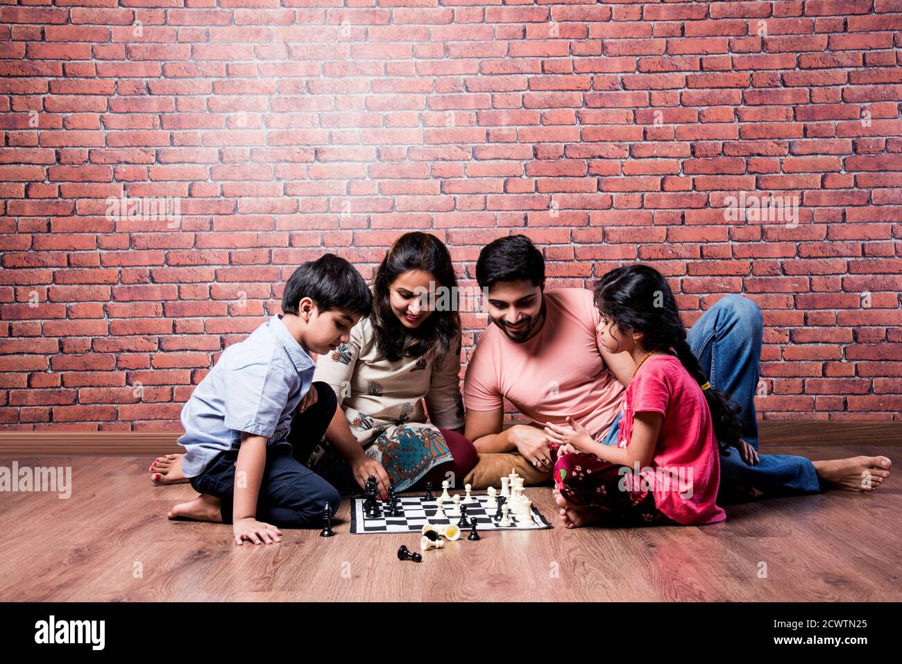 Indian Young Family Of Four Playing Board Games Like Chess Ludo Or Snack And Ladder At Home In indian-young-family-of-four-playing-board-games-like-chess-ludo-or-snack-and-ladder-at-home-in