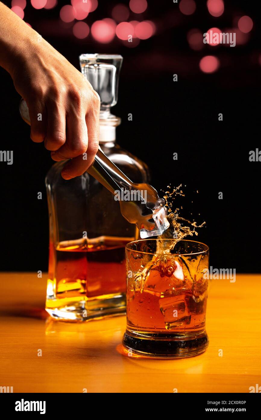 Hand putting ice in a whiskey glass next to a bottle and black ...