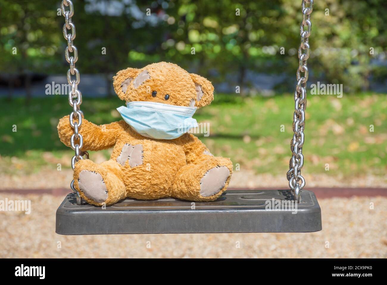 Lonely little teddy bear wearing a medical mask, standing on a swing in ...