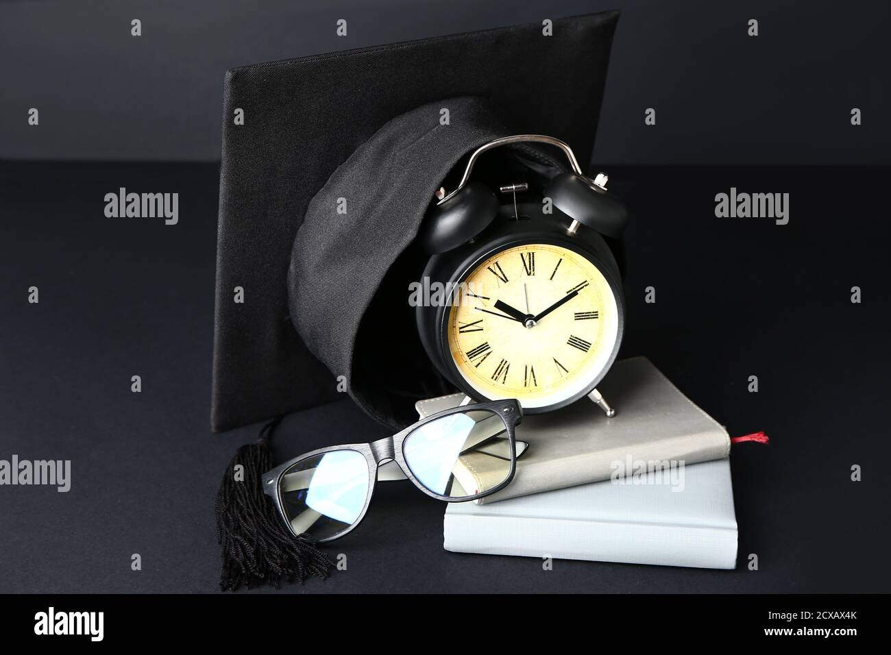 Graduation hat, alarm clock, eyeglasses and books on dark background ...